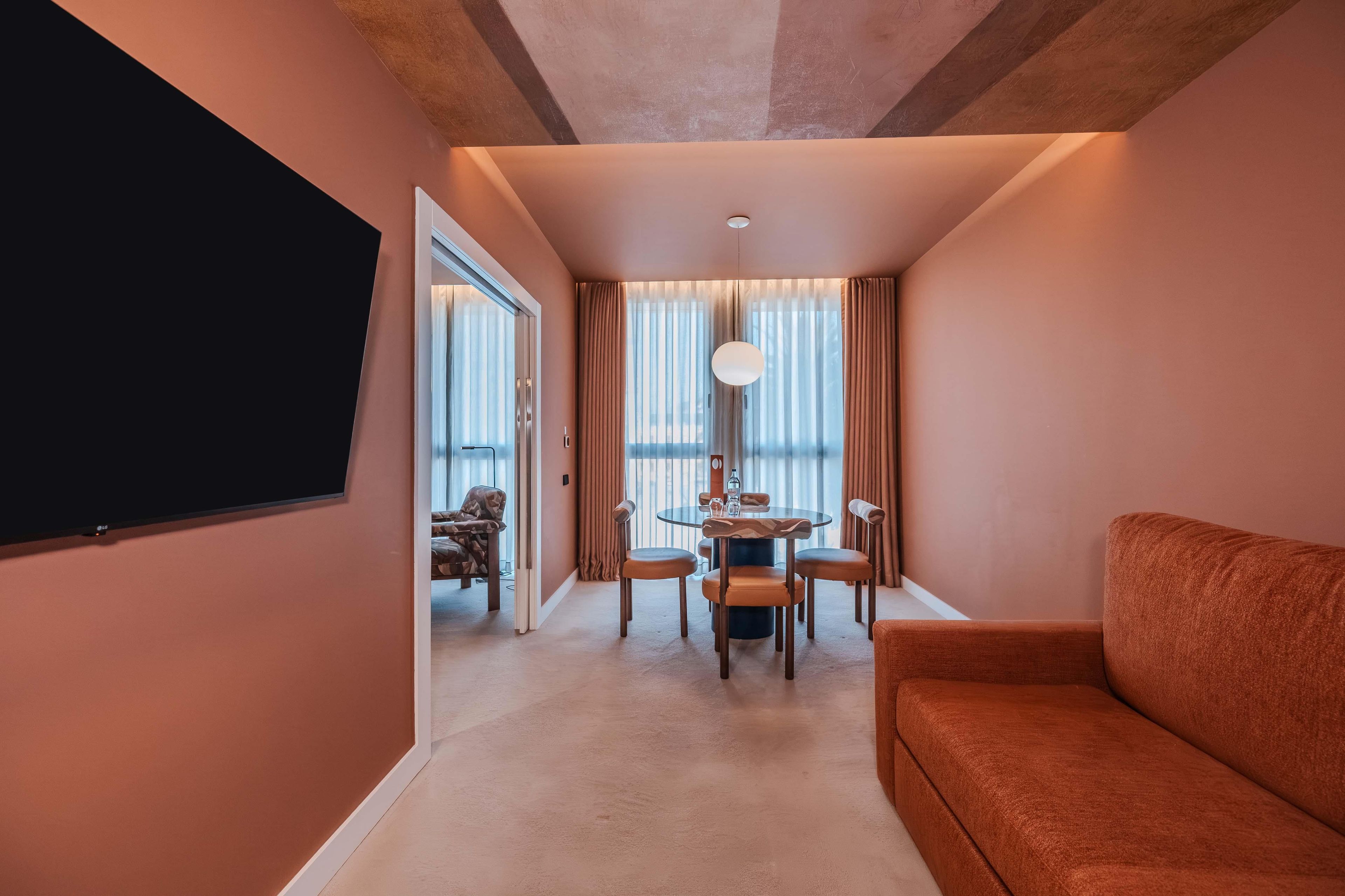Quarto de hotel com televisão de tela plana, mesa de jantar redonda com cadeiras, sofá e uma janela grande com cortinas.