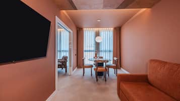 Quarto de hotel com televisão de tela plana, mesa de jantar redonda com cadeiras, sofá e uma janela grande com cortinas.