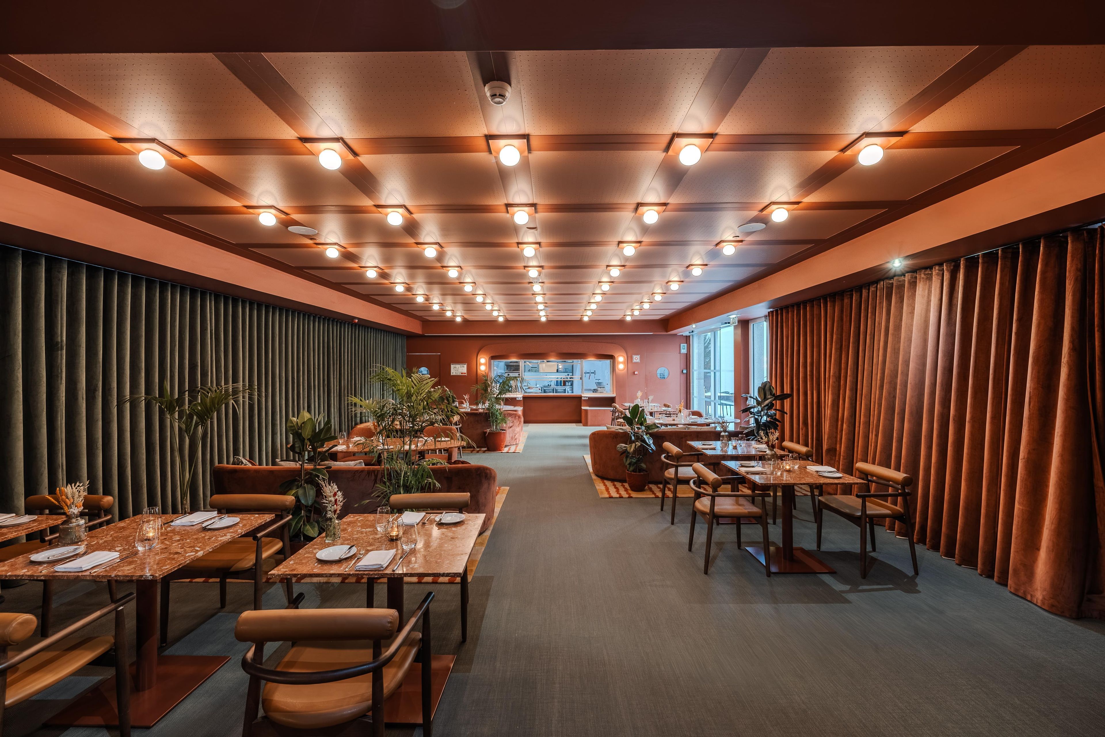 Restaurante com várias mesas e cabines, teto com iluminação embutida e vista para a rua através de grandes janelas.