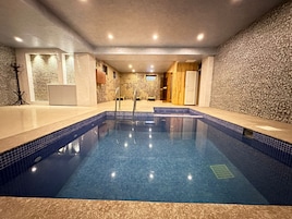 Uma piscina interior com azulejos azuis translúcidos, uma parede de pedra de um lado e uma porta de madeira ao fundo.