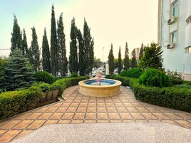 Um pátio com uma fonte central, rodeado por sebes bem aparadas e árvores altas.