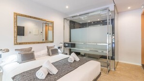 Quarto de hotel moderno com uma cama grande, divisória de vidro, espelho e mesa de cabeceira.