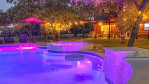 Piscina com borda de pedra e banheira de hidromassagem, cercada por espreguiçadeiras e uma área de estar coberta.
