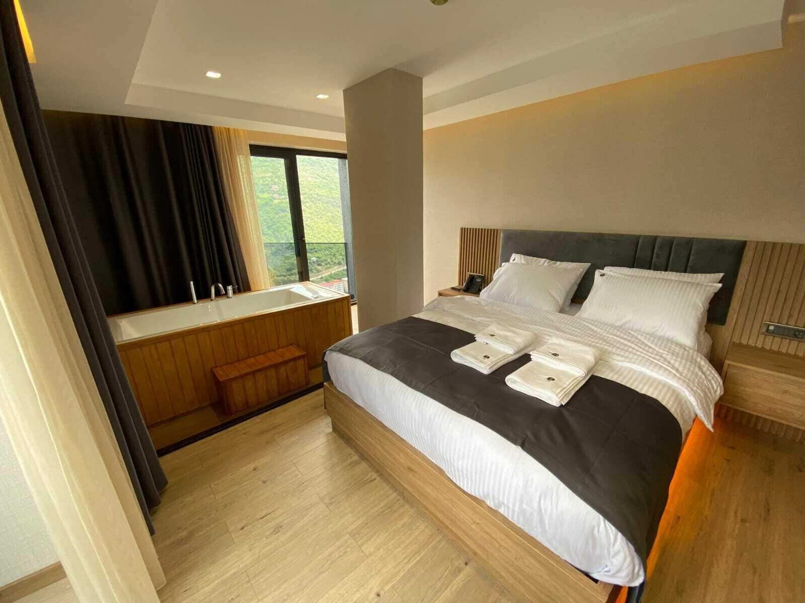 Quarto de hotel moderno com uma cama grande, um banco de madeira, uma janela com cortinas e vista para o verde lá fora.