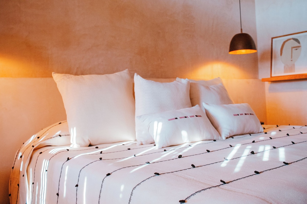 Uma cama bem arrumada com roupa de cama branca e várias almofadas, uma luminária pendente acima e um quadro na parede.