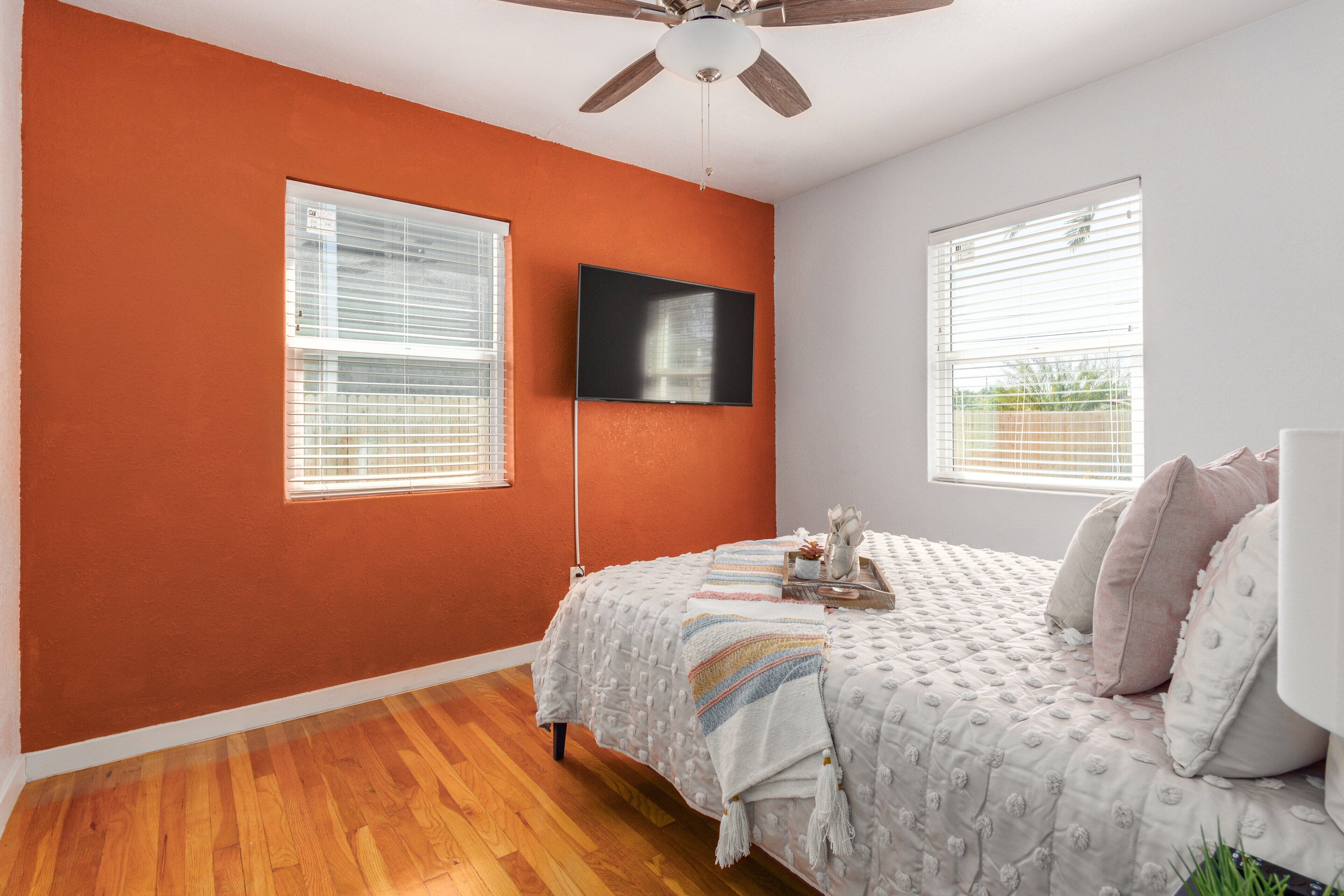 Quarto com parede laranja, piso de madeira, cama com colcha branca e estampada, duas janelas com persianas e um ventilador de teto.