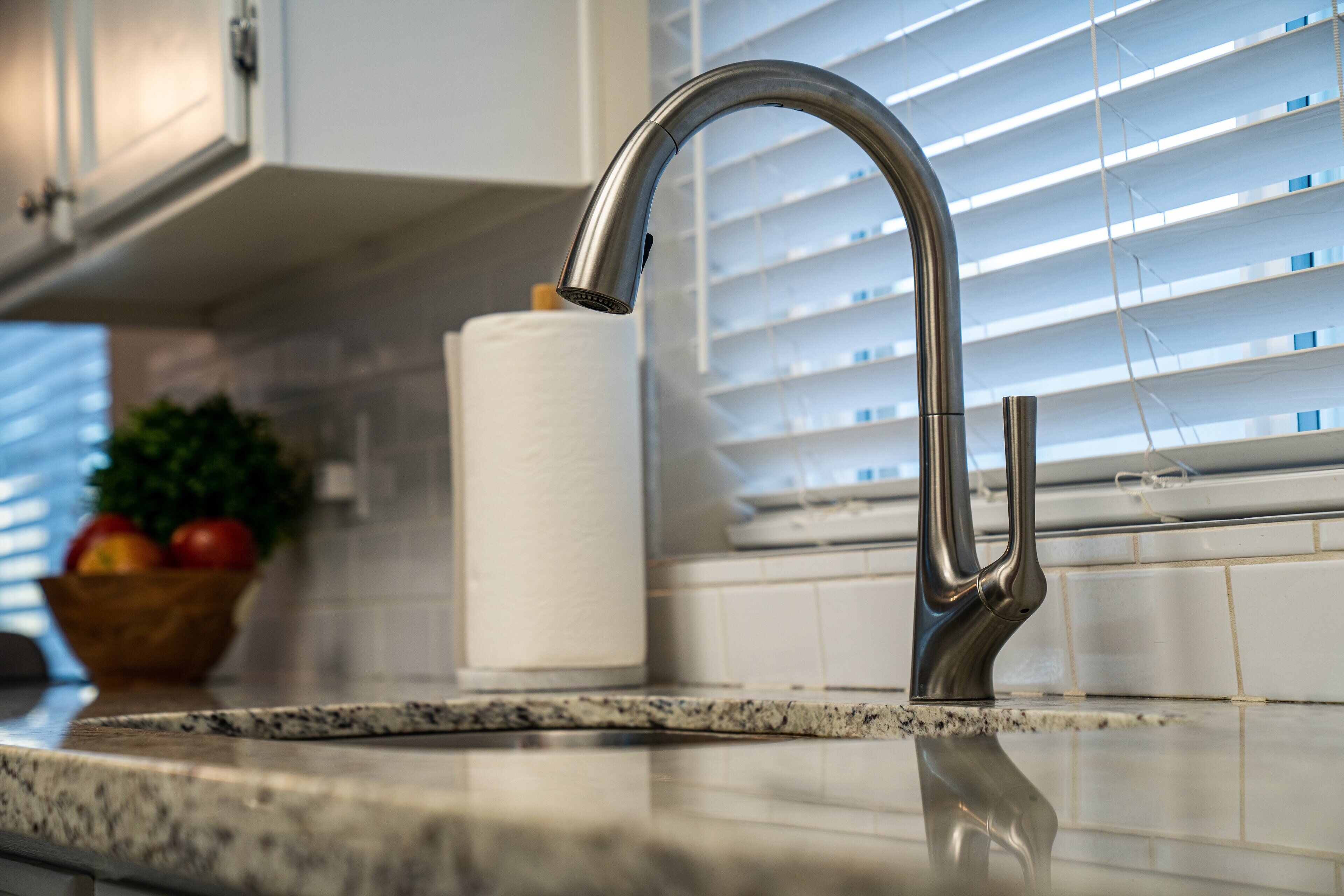 Uma cozinha com uma torneira de aço inoxidável, um suporte de papel toalha branco e uma tigela de frutas no balcão.