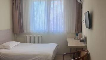 Quarto de hotel com cama, escrivaninha, cadeira, televisão e janela com cortinas.