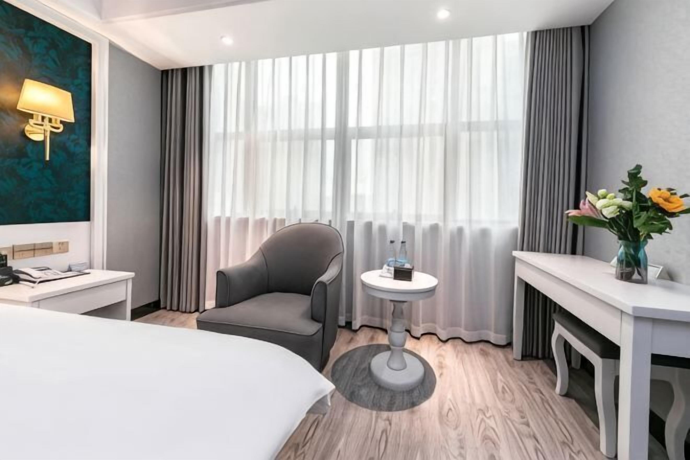 Quarto de hotel com cama, escrivaninha, cadeira, mesa pequena e janela com cortinas.