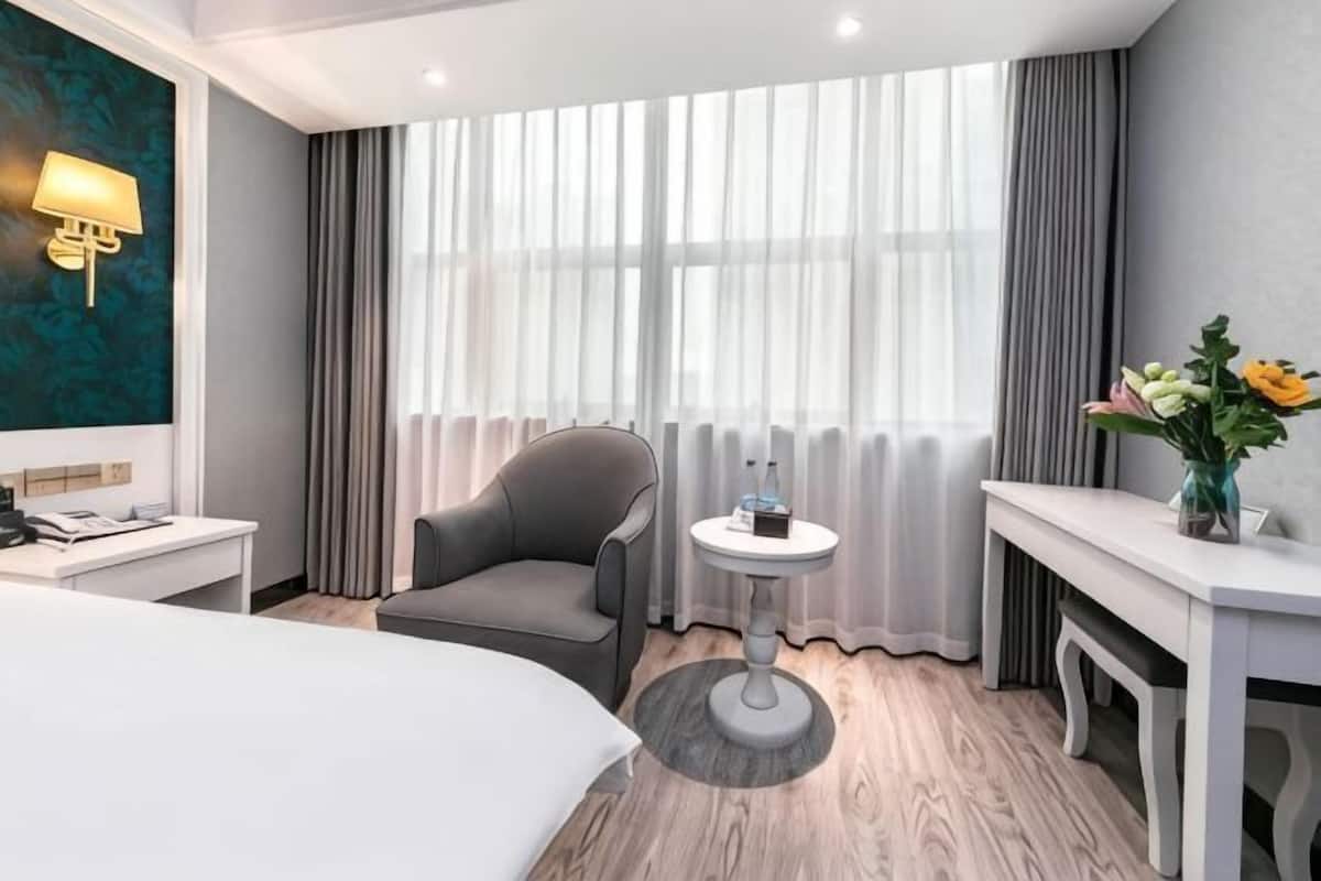 Quarto de hotel com cama, escrivaninha, cadeira, mesa pequena e janela com cortinas.