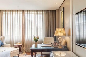 Quarto de hotel com cama, escrivaninha com luminária, cadeira, pequena mesa com um vaso de flores e uma janela grande com cortinas.