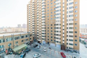Um edifício residencial alto com vários andares, área de estacionamento na frente e vista para a paisagem urbana ao redor.