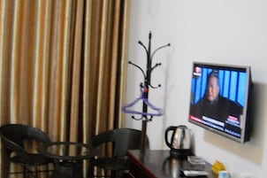 Quarto com televisão na parede, mesa de centro, duas cadeiras e um cabideiro.