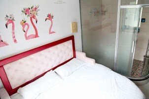 Quarto com cabeceira vermelha, roupa de cama branca, box de chuveiro envidraçado e parede com arte floral.