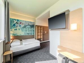 Quarto de hotel moderno com cama, televisão de tela plana, mesa de cabeceira de madeira, um banquinho pequeno de madeira e prateleira fixada na parede.