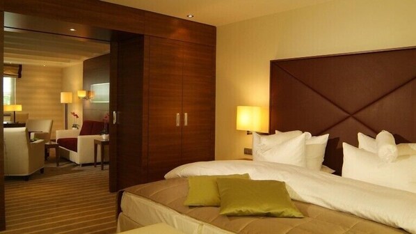 Quarto de hotel com uma cama grande, um guarda-roupa de madeira, uma escrivaninha com cadeira e uma pequena área de estar com uma cadeira vermelha e uma mesa.