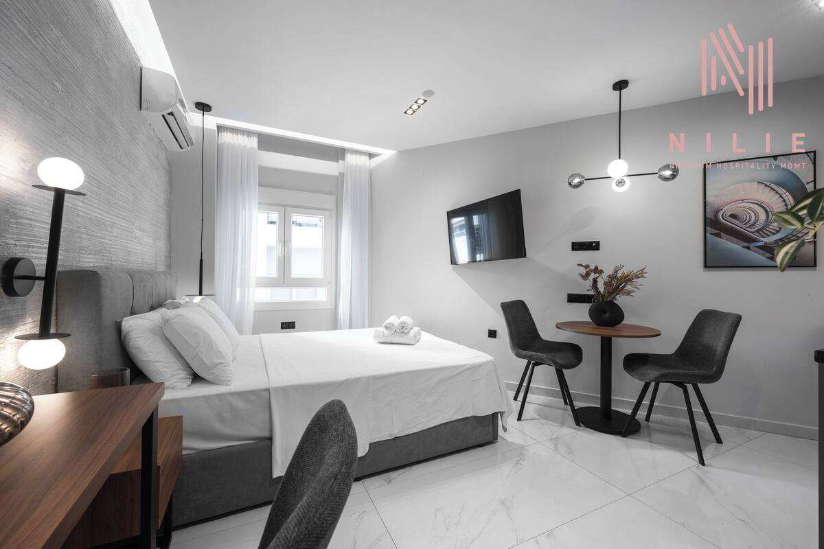 Quarto de hotel moderno com cama, mesa de cabeceira, abajur, TV e uma pequena área de refeições com cadeiras e mesa.