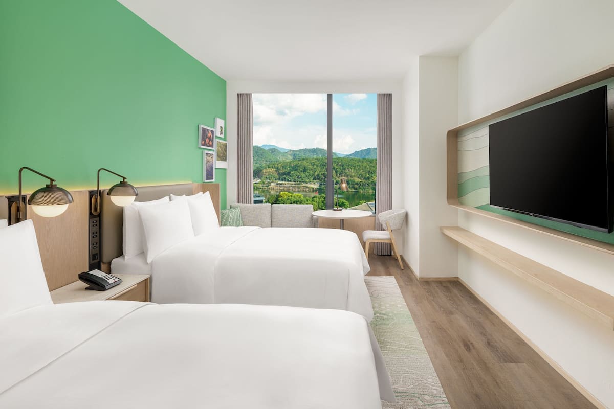 Quarto de hotel com duas camas, uma televisão grande e vista para montanhas e um lago.