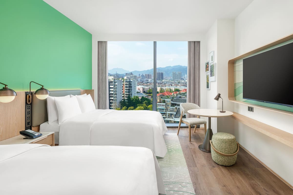Quarto de hotel com duas camas, uma mesa pequena, uma cadeira, televisão e vista para a cidade e montanhas.