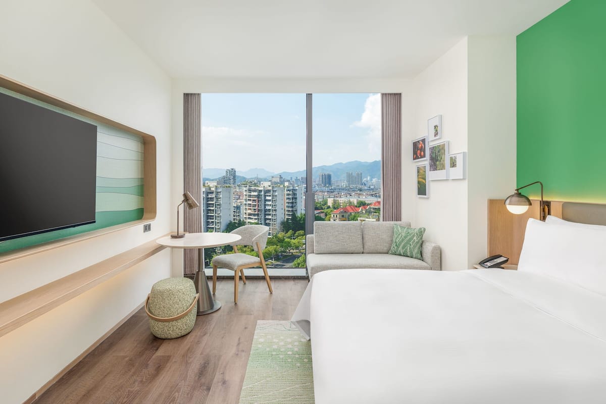Quarto de hotel moderno com uma cama grande, uma mesinha redonda pequena, um sofá e uma televisão fixada na parede.