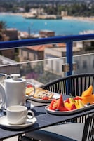Mesa na varanda com cafeteira, xícara e prato de frutas e doces, com vista para a praia e edifícios.