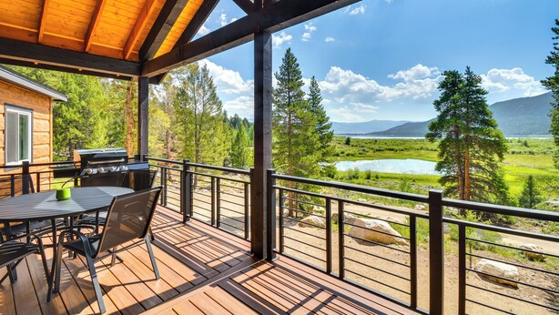 Um deck de madeira com mesa e cadeiras, com vista para um lago e montanhas arborizadas.