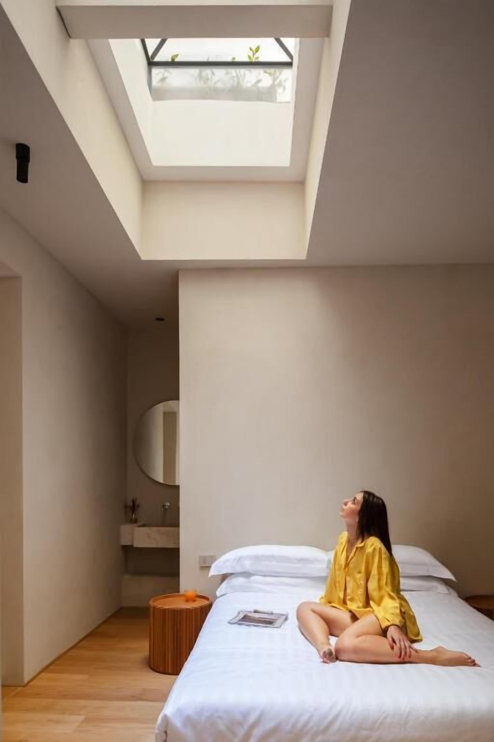 Quarto com claraboia, cama, espelho redondo e um banquinho de madeira.