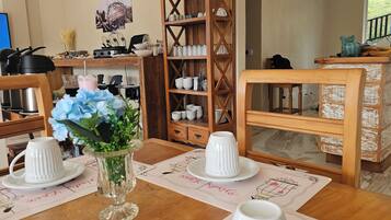 Uma área de refeições com uma mesa de madeira posta para dois, um vaso com flores azuis e uma cozinha ao fundo.