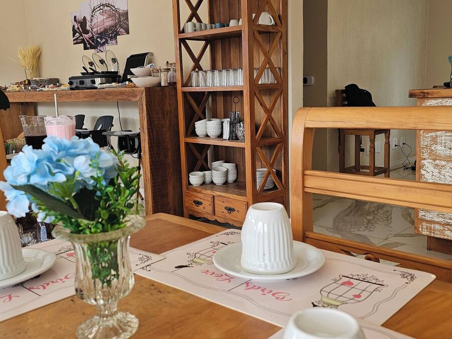 Uma área de refeições com uma mesa de madeira posta para dois, um vaso com flores azuis e uma cozinha ao fundo.