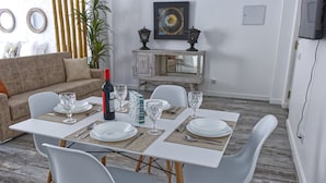 Uma área de refeições com uma mesa branca posta para quatro pessoas, rodeada por cadeiras brancas. Há um sofá e um armário ao fundo.