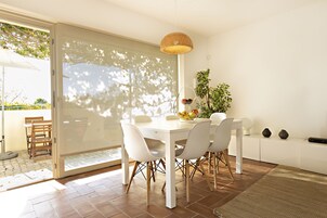 Uma sala de jantar com mesa e cadeiras brancas, uma luminária pendente trançada e uma planta ao lado.