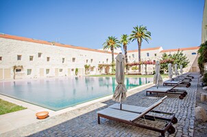 Uma área à beira da piscina com espreguiçadeiras e guarda-sóis, piscina translúcida e um edifício com telhado em azulejos ao fundo.