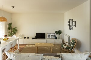 Uma sala de estar moderna com um sofá branco, uma mesa de centro de madeira, uma TV sobre um móvel branco e uma cadeira de vime.