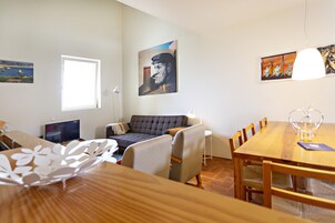 Uma sala de estar moderna com um sofá, uma mesa de jantar e cadeiras. Há um quadro na parede e uma janela que proporciona luz natural.