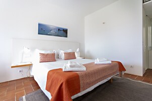 Quarto de hotel bem organizado com uma cama grande, uma mesinha de cabeceira, um quadro na parede e vista para o mar pela janela.