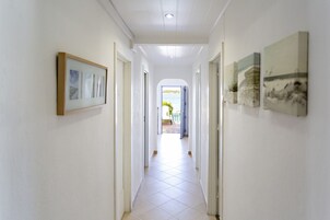 Um corredor com paredes brancas, obras de arte emolduradas e um chão de azulejos que leva a uma porta com vista para o exterior.