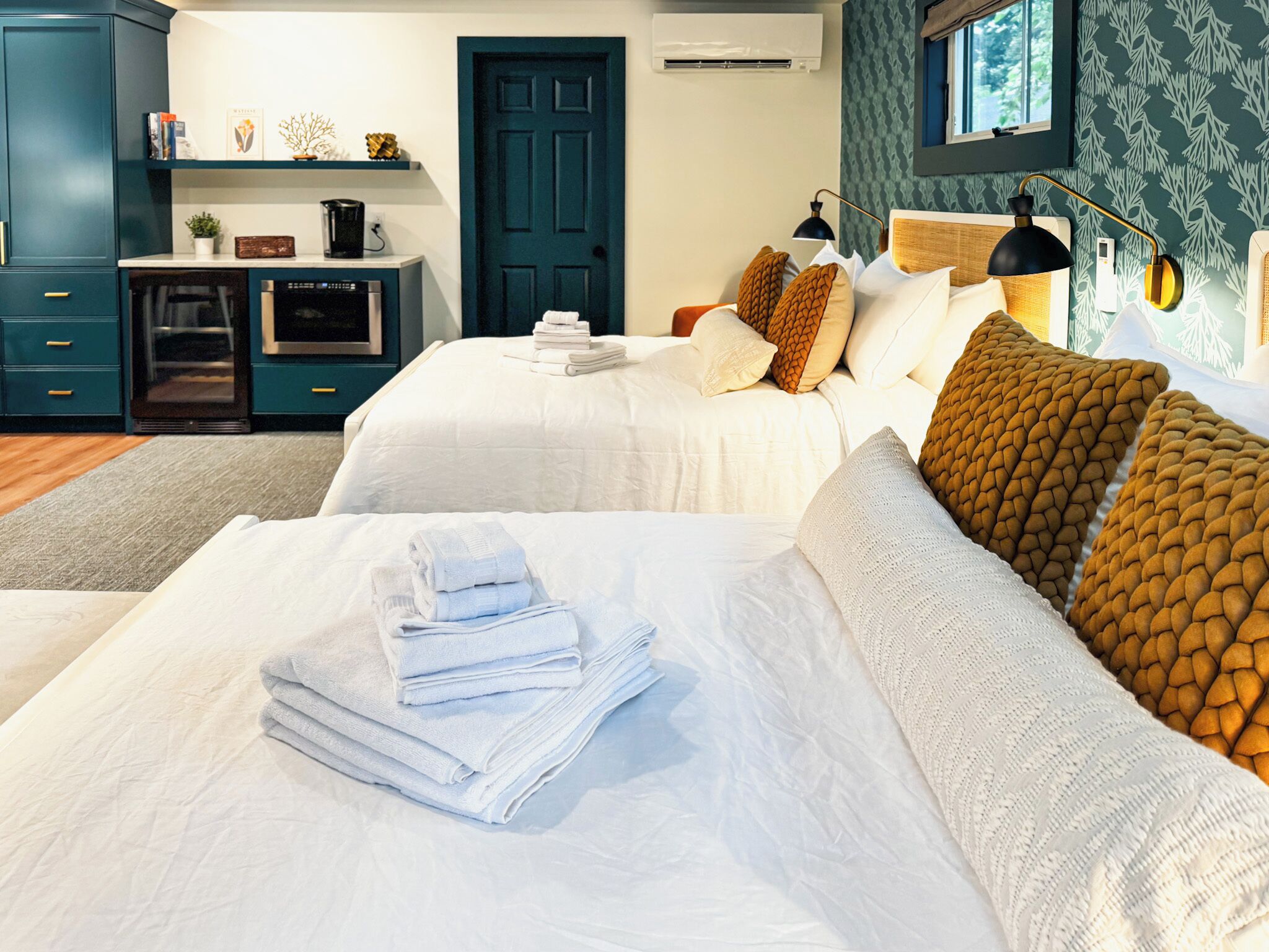 Quarto de hotel com duas camas, uma cozinha compacta, uma porta azul e uma janela com vista.
