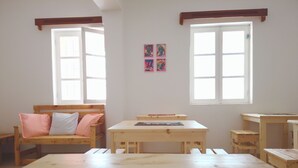 Quarto com uma mesa de madeira, um banco de madeira com almofadas, duas janelas e uma parede com obras de arte emolduradas.