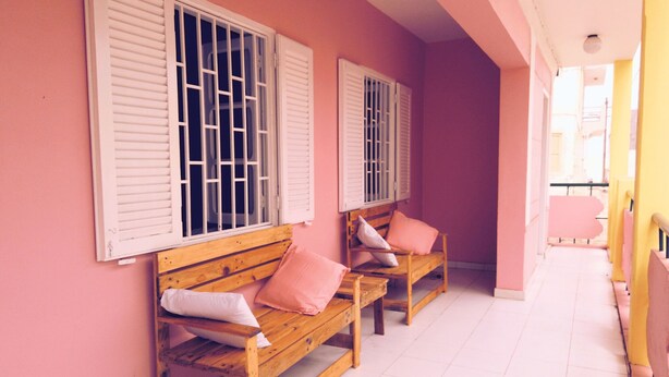 Um corredor com duas bancas de madeira encostadas a paredes cor-de-rosa, cada uma com uma janela e uma persiana branca.