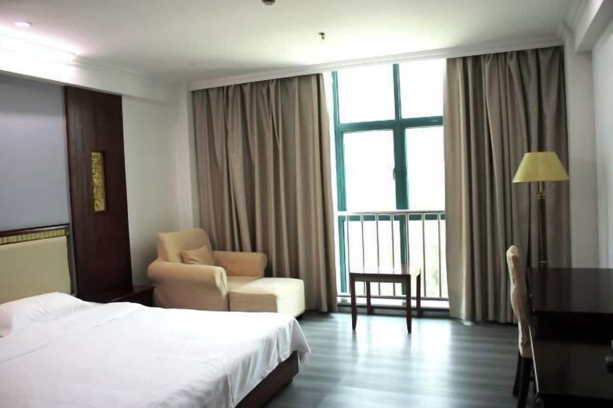Quarto de hotel com cama, poltrona, escrivaninha, luminária e janela com cortinas.