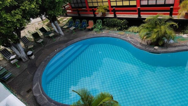 Piscina curva com padrão quadriculado, cercada por cadeiras de descanso e plantas em vasos.