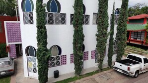 Um edifício branco com várias janelas e painéis decorativos, uma caminhonete branca estacionada na frente e um ônibus vermelho ao fundo.