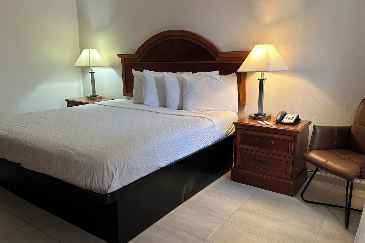 Uma cama bem arrumada com cabeceira de madeira, duas mesas de cabeceira com abajures, uma poltrona de couro marrom e uma pequena escrivaninha com um telefone.