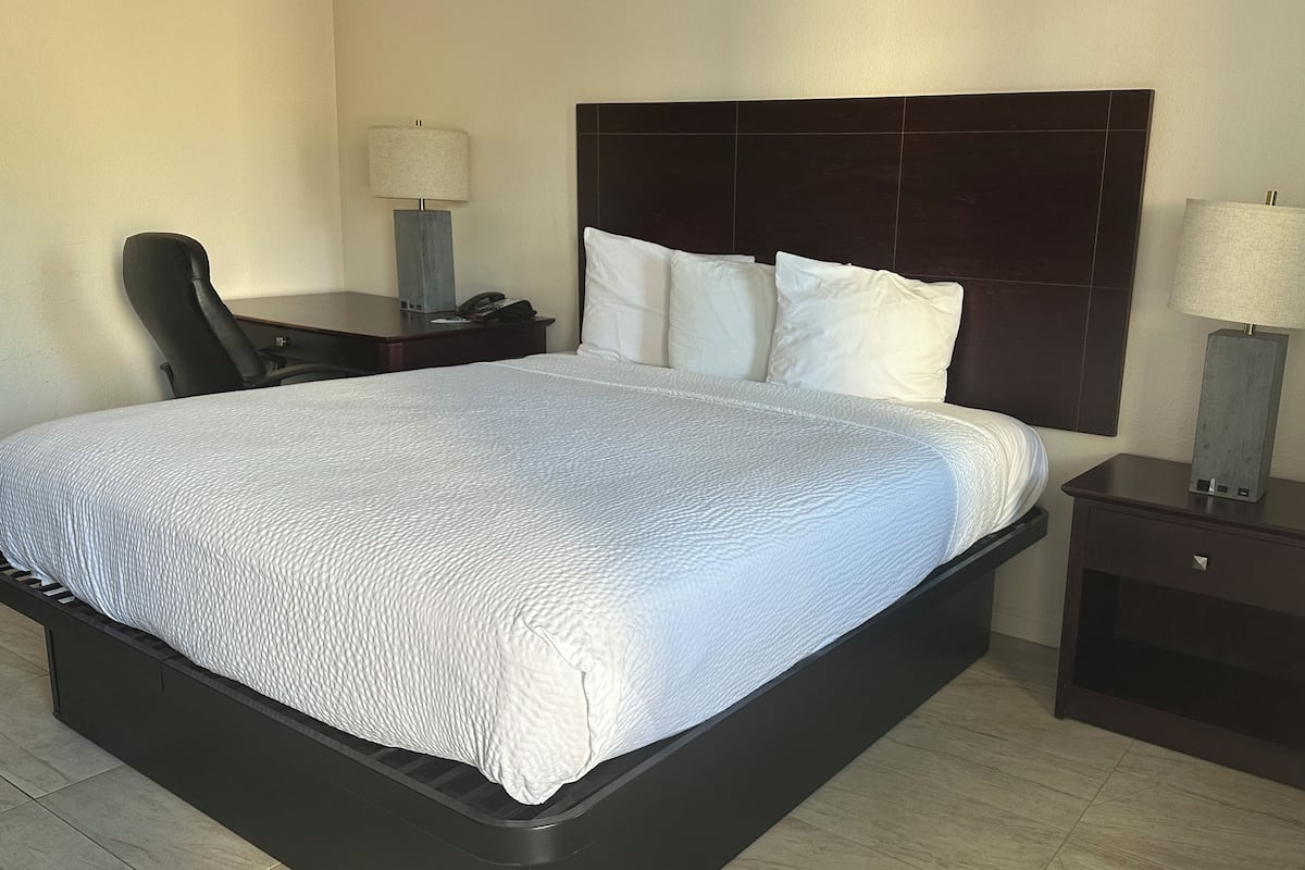 Quarto de hotel com uma cama grande, cabeceira escura, duas mesas de cabeceira com abajures e uma cadeira.