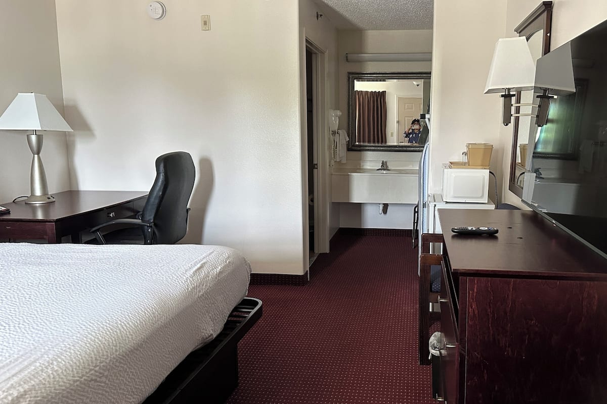 Quarto de hotel com cama, escrivaninha com luminária, cadeira, televisão, cómoda e banheiro visível por uma porta aberta.