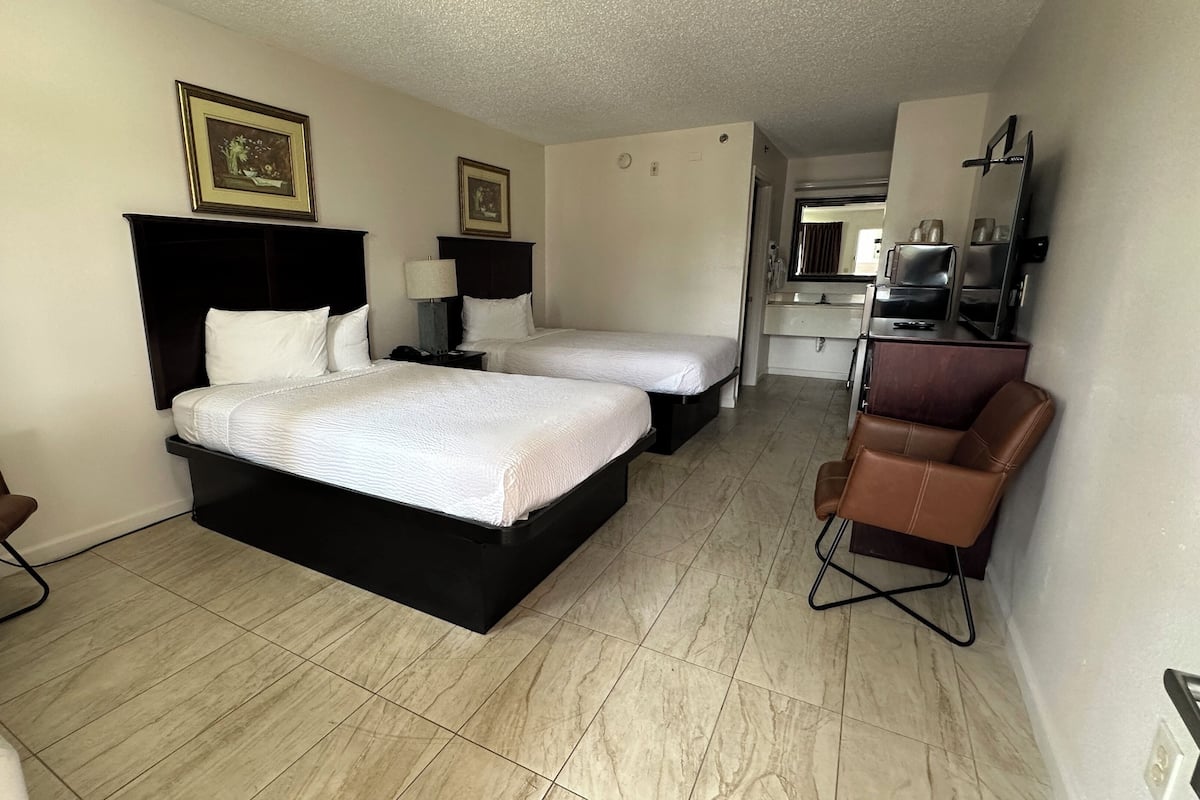 Quarto de hotel com uma cama grande, uma cama menor, uma escrivaninha, uma cadeira e uma cozinha compacta.