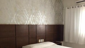 Quarto de hotel com uma cama grande, uma escrivaninha e uma cadeira. Há um ar condicionado fixado na parede e uma cortina na janela.