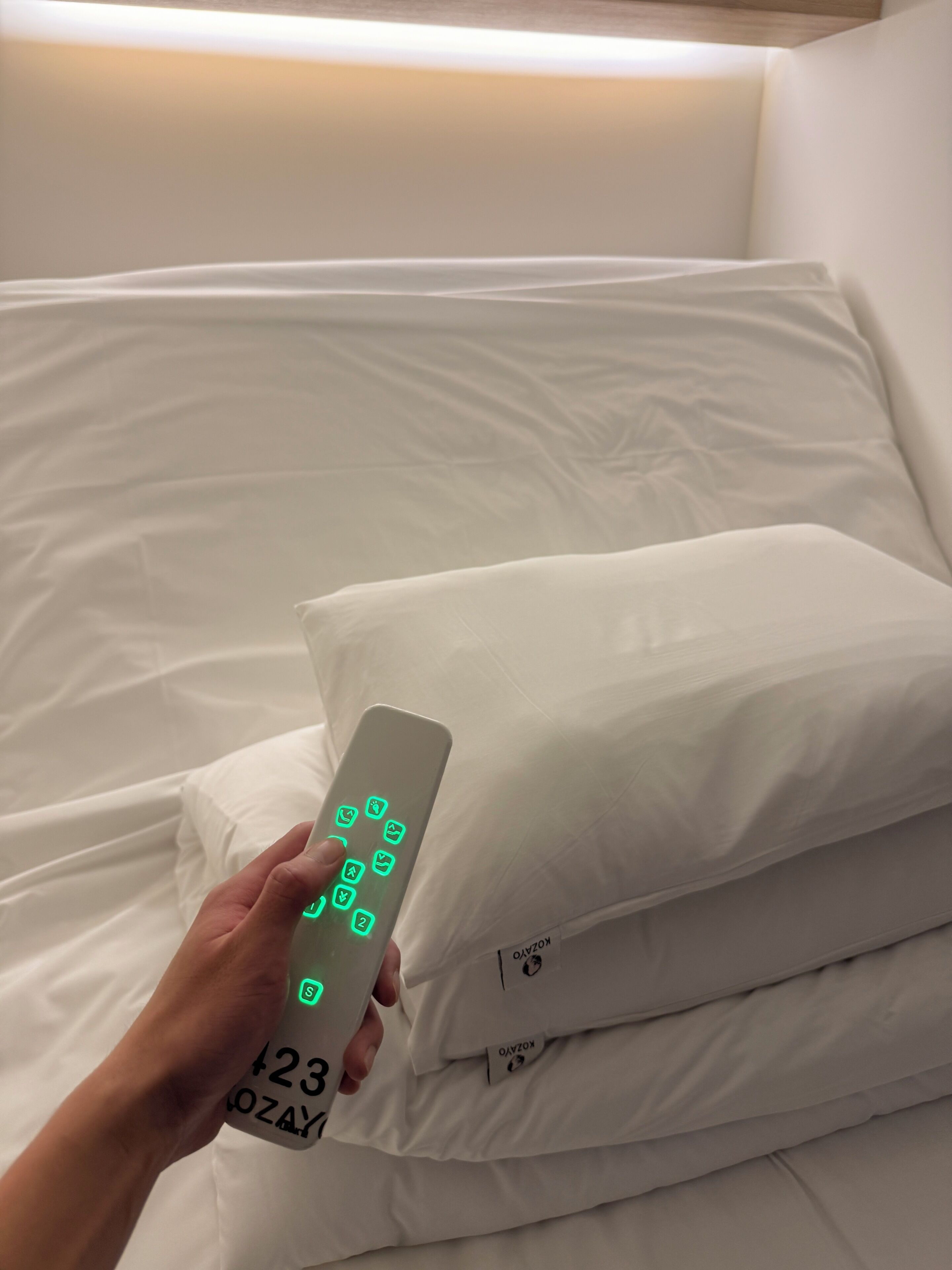 Mão segurando um controle remoto com botões iluminados, apoiado em uma cama bem arrumada com roupa de cama e travesseiros brancos.