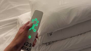 Mão segurando um controle remoto com botões iluminados, apoiado em uma cama bem arrumada com roupa de cama e travesseiros brancos.