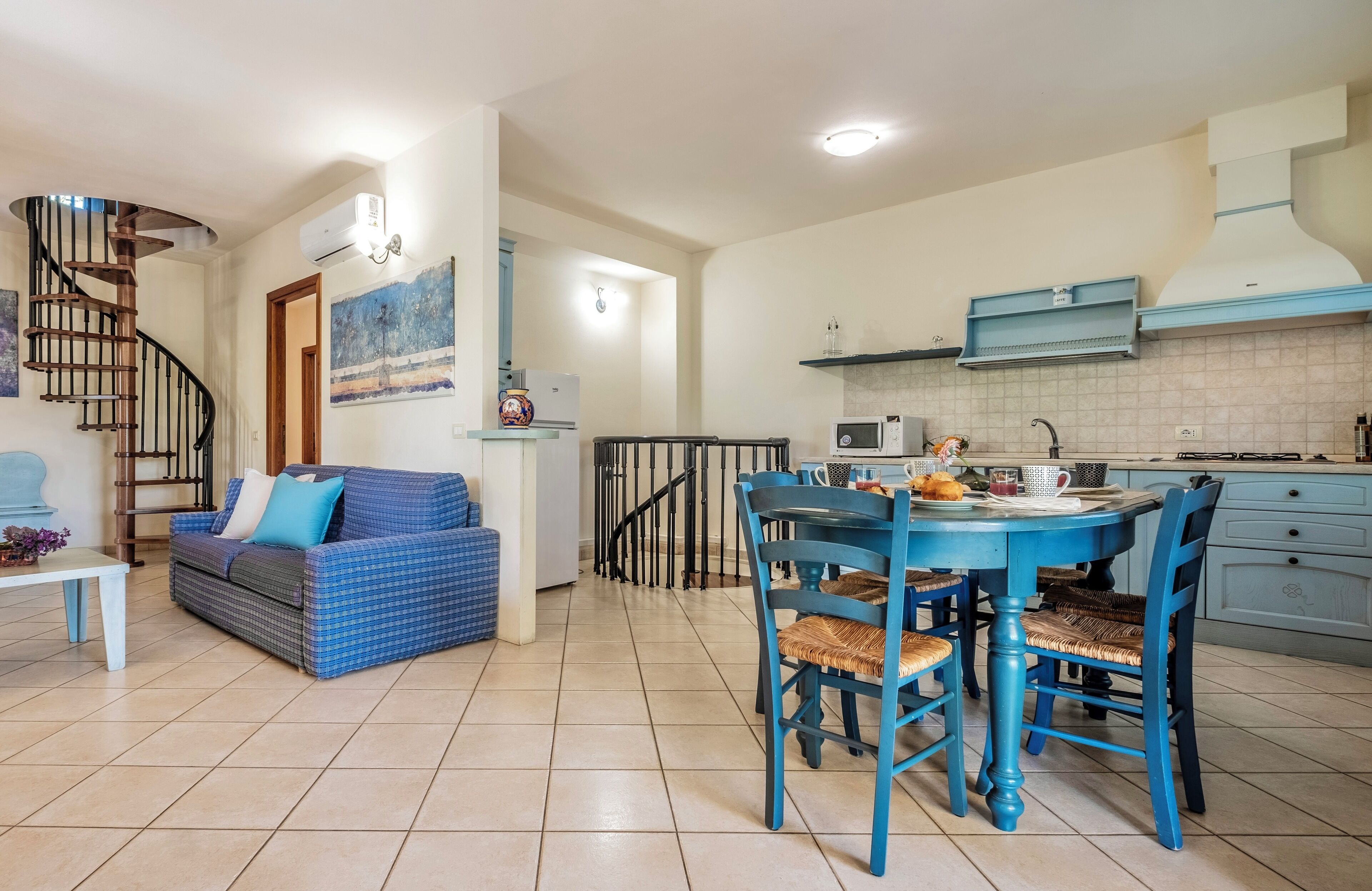 Cozinha moderna com área de refeições, destacando uma escada em espiral, um sofá azul e uma mesa de jantar com cadeiras.